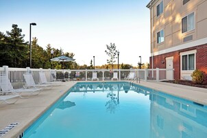 Piscina com cadeiras de descanso e guarda-sóis, cercada por um muro branco e árvores.