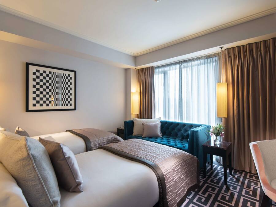 Quarto de hotel com uma cama grande, um sofá menor, uma cadeira, uma luminária e uma janela com cortinas.