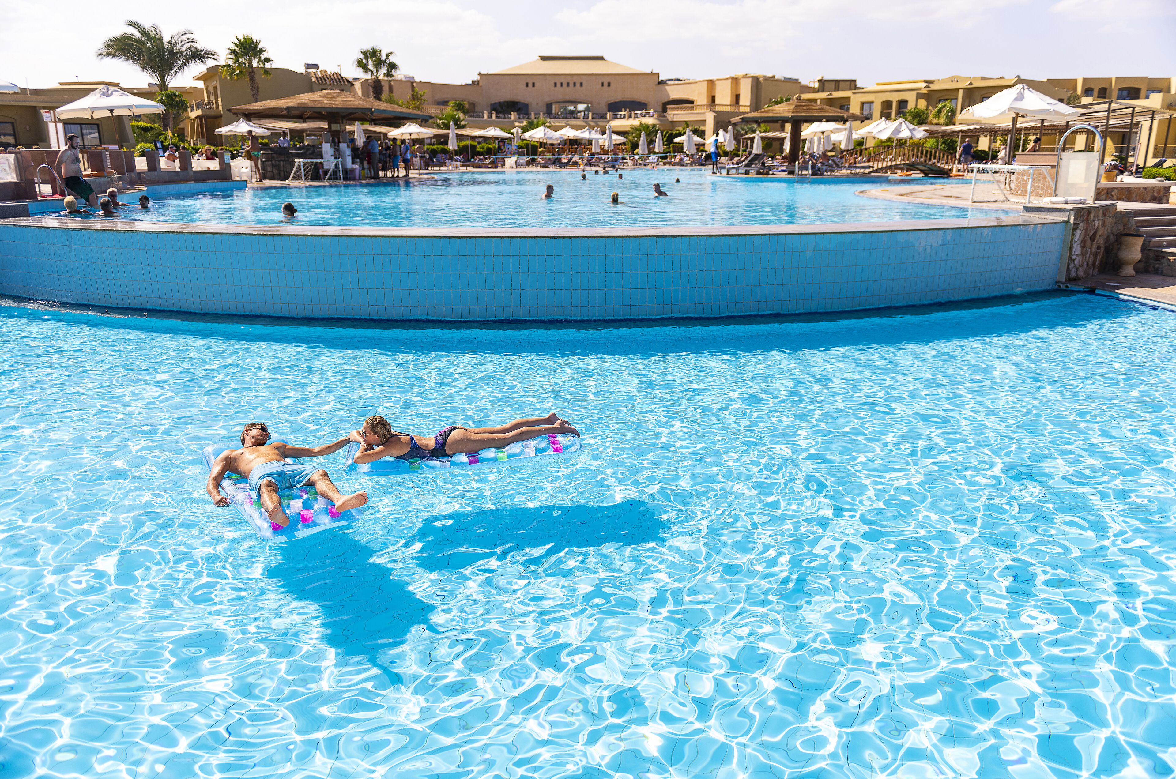 Uma piscina grande com pessoas relaxando em bóias, cercada por guarda-sóis e espreguiçadeiras.