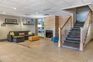 Uma sala de estar moderna com sofá, banquinho e lareira. Uma escada com corrimão de madeira leva a um andar superior.