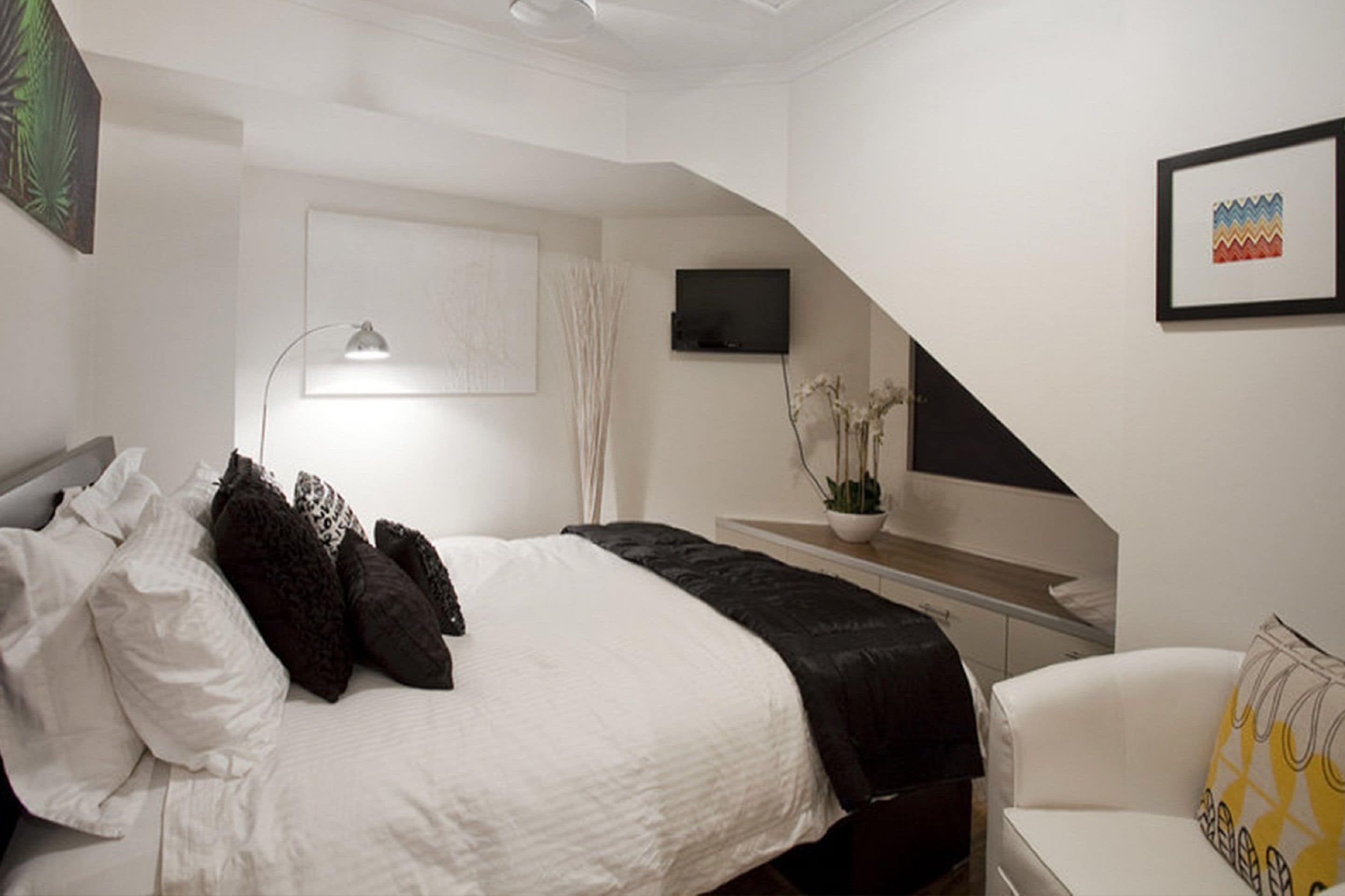 Quarto moderno com cama, escrivaninha e cadeira. Há uma TV fixada na parede e uma planta na escrivaninha.