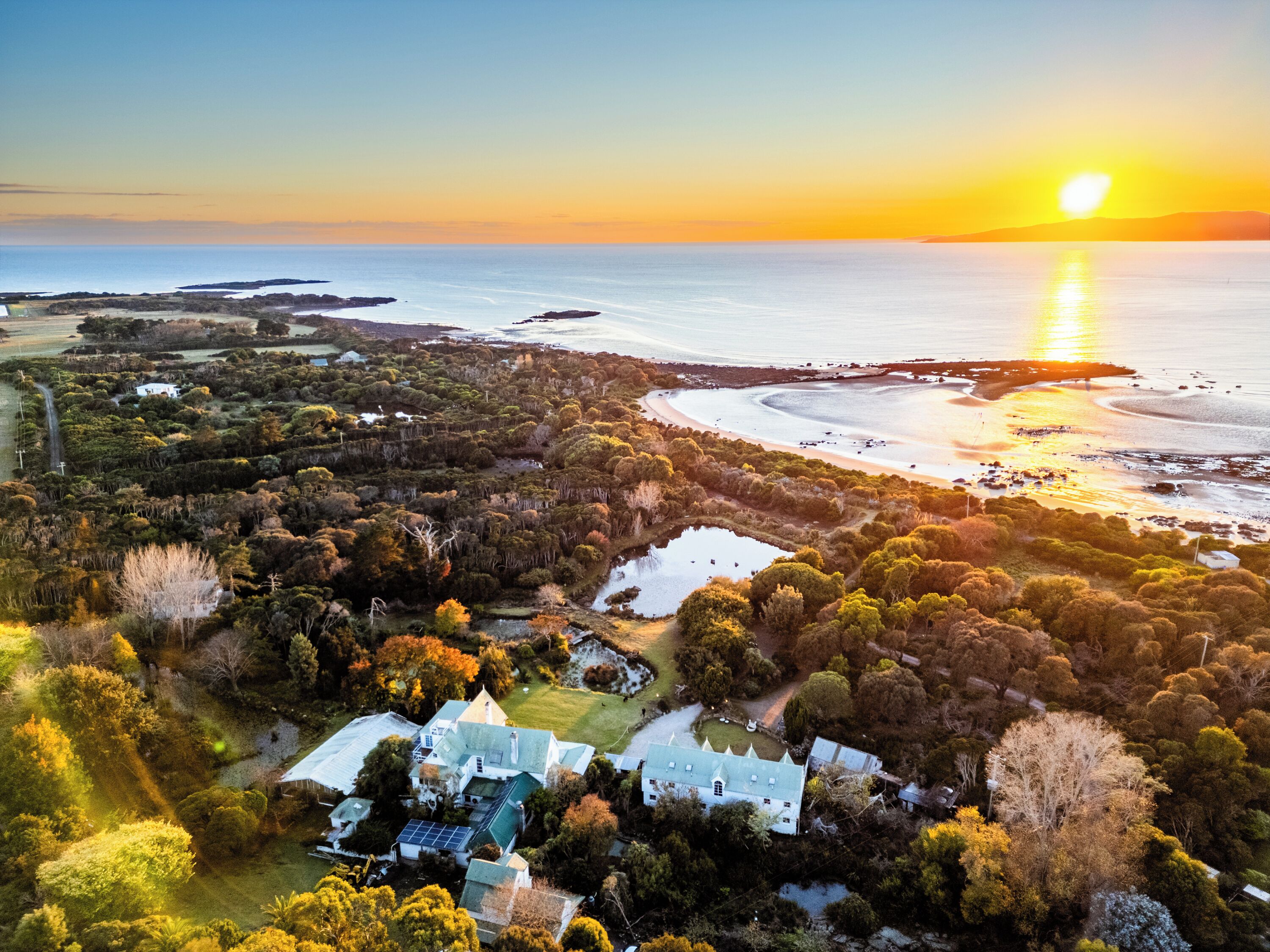 Vista aérea de uma propriedade costeira com uma casa grande, rodeada por vegetação densa e uma praia.