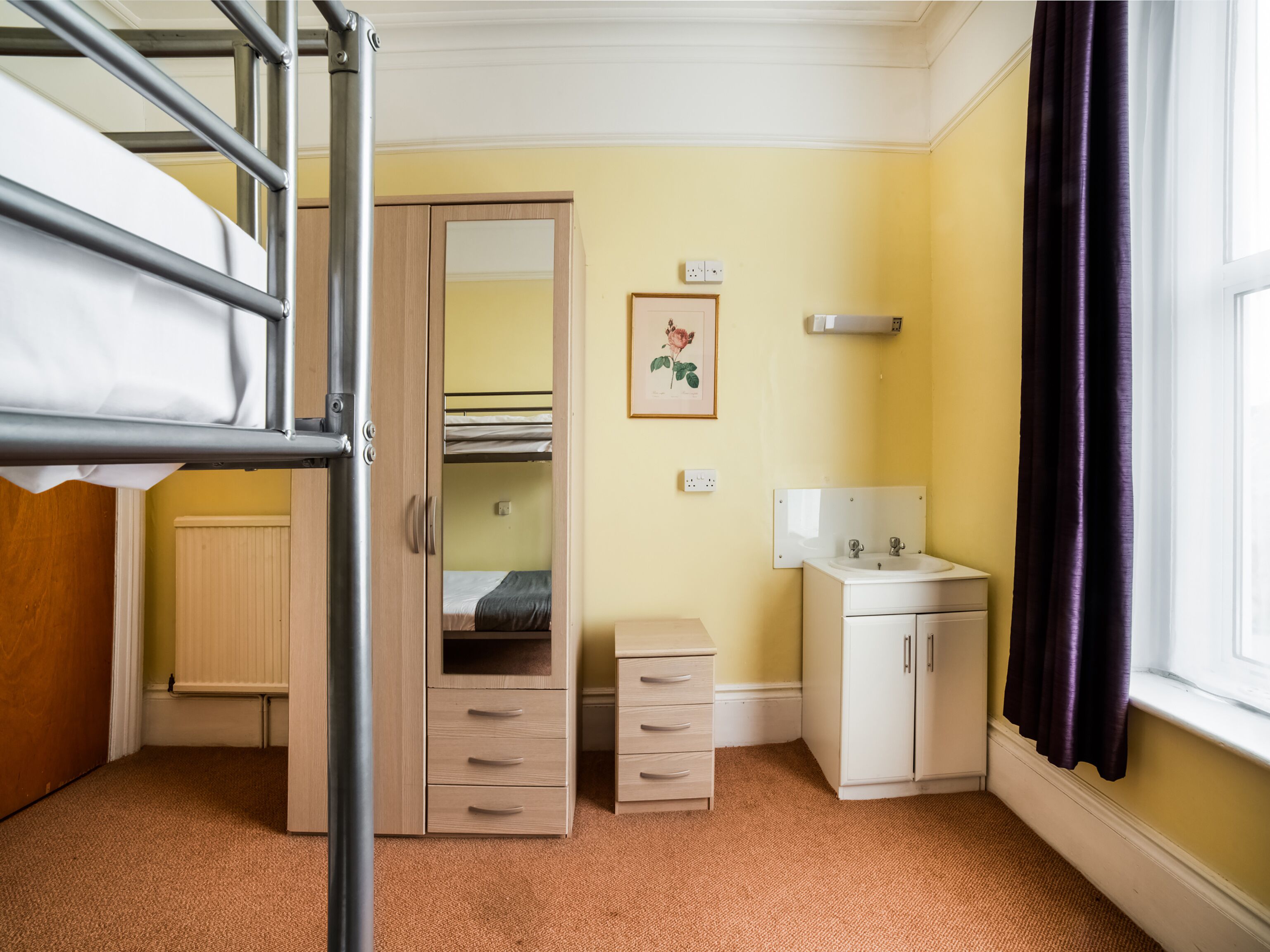 Quarto compacto com beliche, pequeno armário, pia e janela com cortinas.
