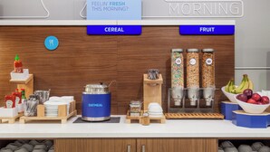 Buffet de café da manhã com seções identificadas para cereais e frutas, incluindo um recipiente azul para mingau e potes de cereais.