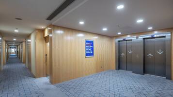 Corredor de hotel com paredes revestidas de madeira, carpete estampado e um elevador com setas indicativas.