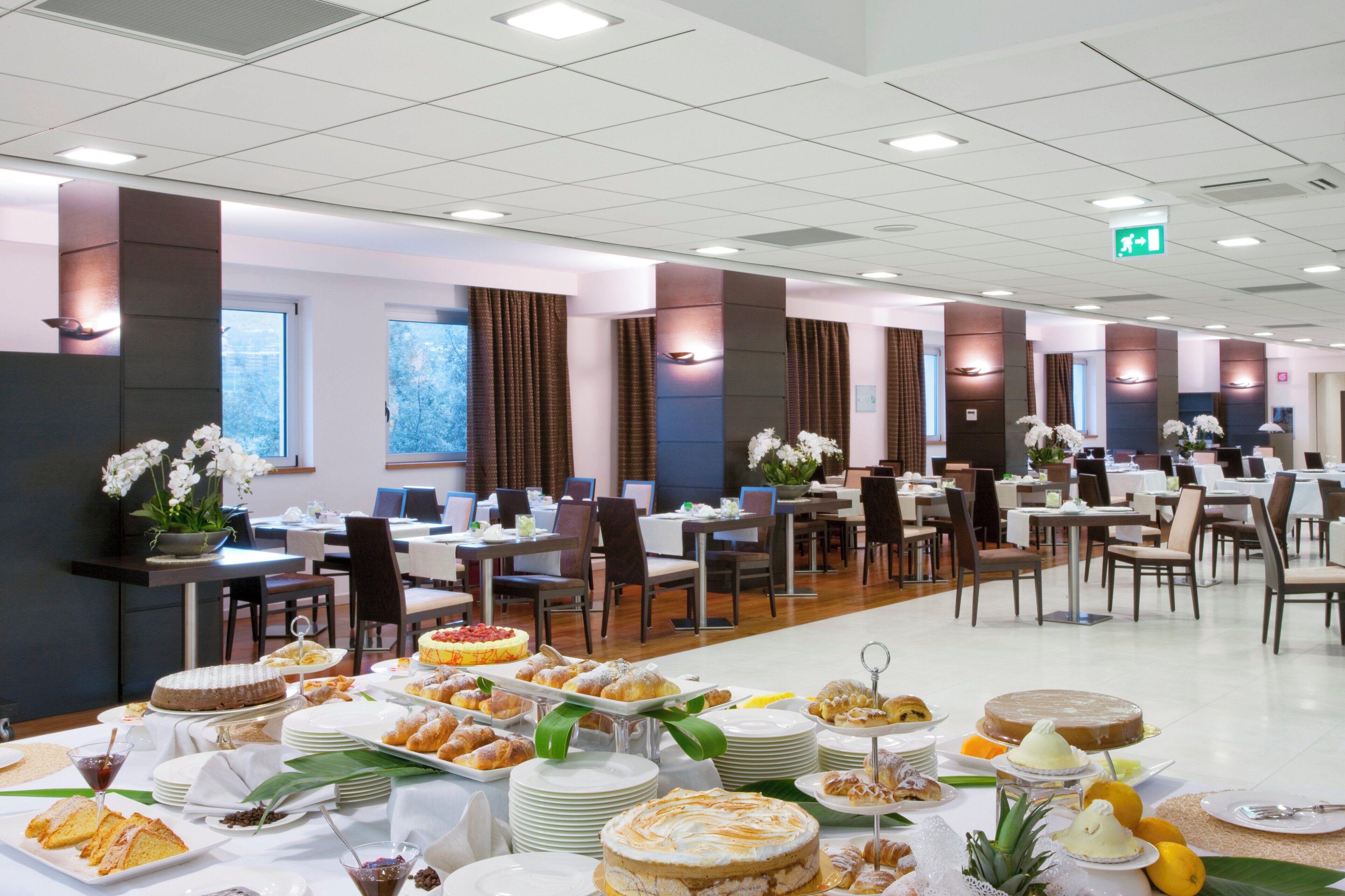 Espaço de refeições de hotel com várias mesas, cadeiras, sobremesas e doces.