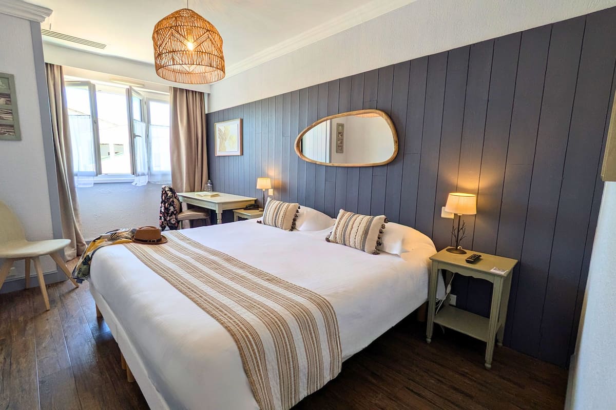 Quarto de hotel com uma cama grande, duas mesas de cabeceira com luminárias, uma cadeira, uma escrivaninha e uma janela ampla com cortinas.
