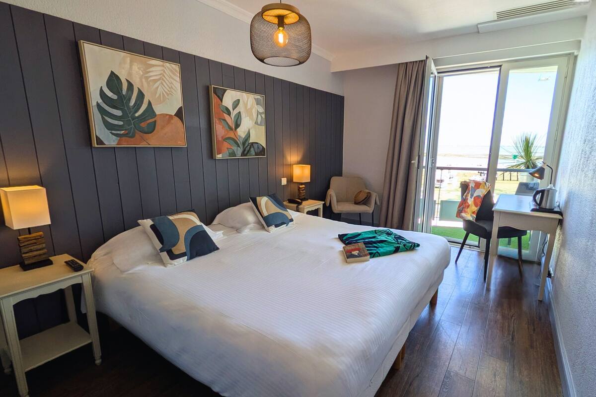 Quarto de hotel com uma cama grande, duas mesas de cabeceira, uma escrivaninha e uma cadeira. Há duas luminárias na parede e uma luz no teto.