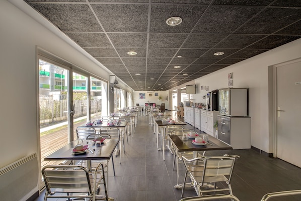 Uma sala de jantar ampla, com mesas e cadeiras, uma área de cozinha com eletrodomésticos, e grandes janelas que proporcionam iluminação natural.