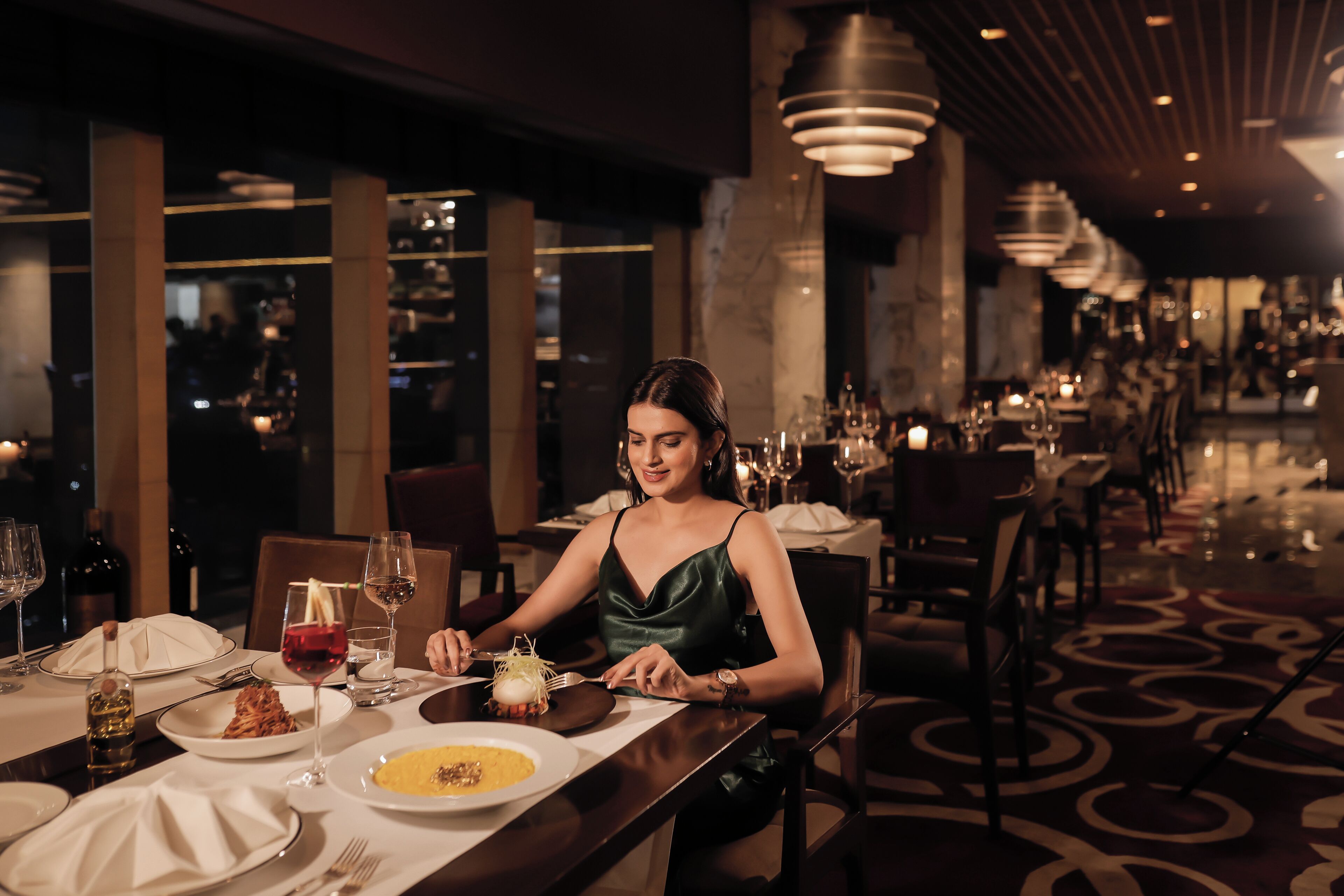 Uma mulher jantando em um restaurante, com uma mesa posta para refeição, incluindo pratos de comida e taças de vinho.