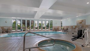 Piscina interna com banheira de hidromassagem, janelas de vidro transparente e equipamentos de ginástica.