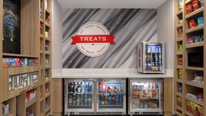 Uma loja com uma placa 'Treats', prateleiras cheias de lanches e bebidas, e uma geladeira com bebidas.