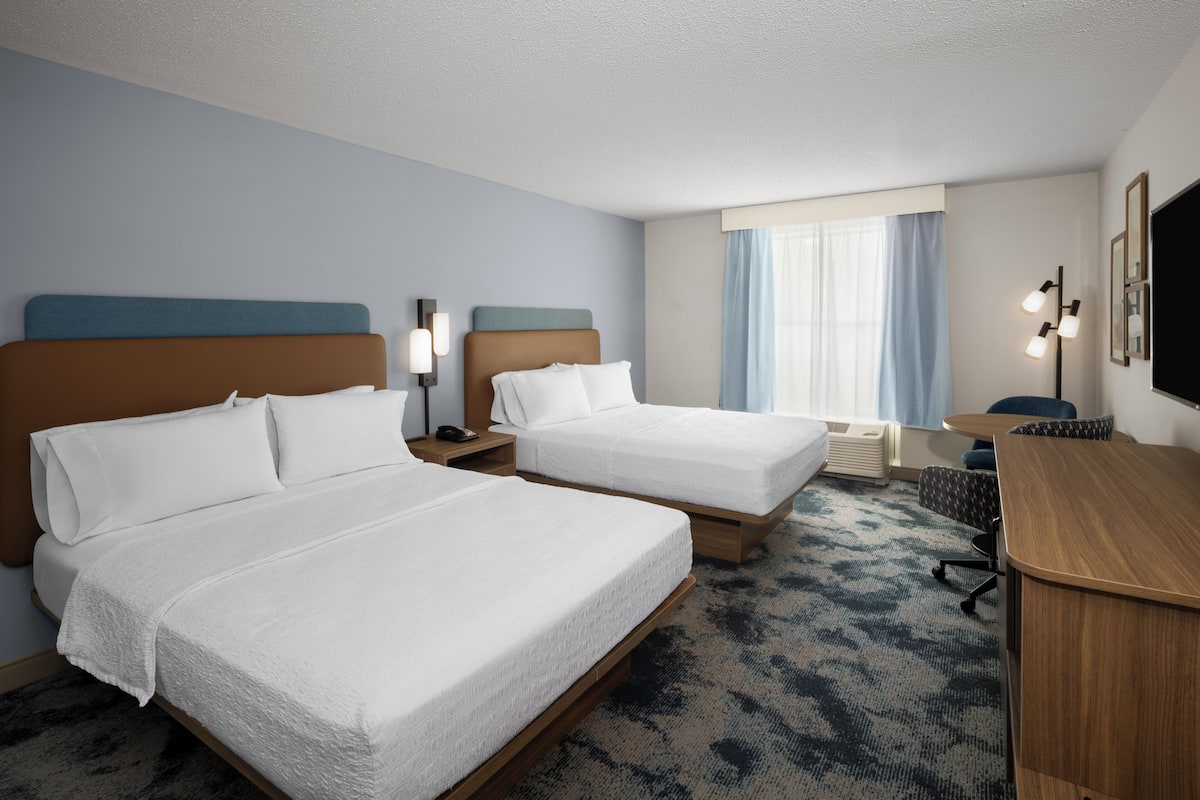 Quarto de hotel com duas camas, uma escrivaninha, uma cadeira, um abajur e uma janela com cortinas.