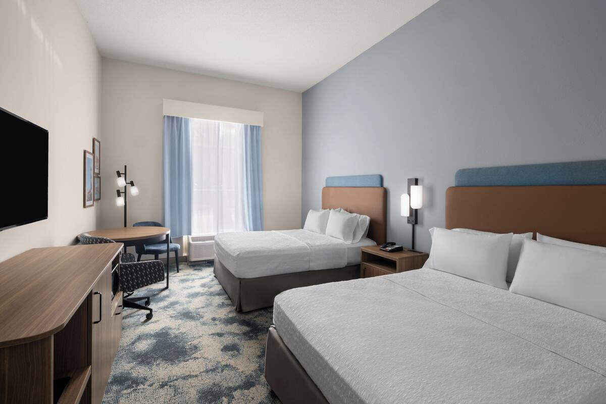 Quarto de hotel com duas camas, uma escrivaninha, uma cadeira, uma televisão e uma janela com cortinas.