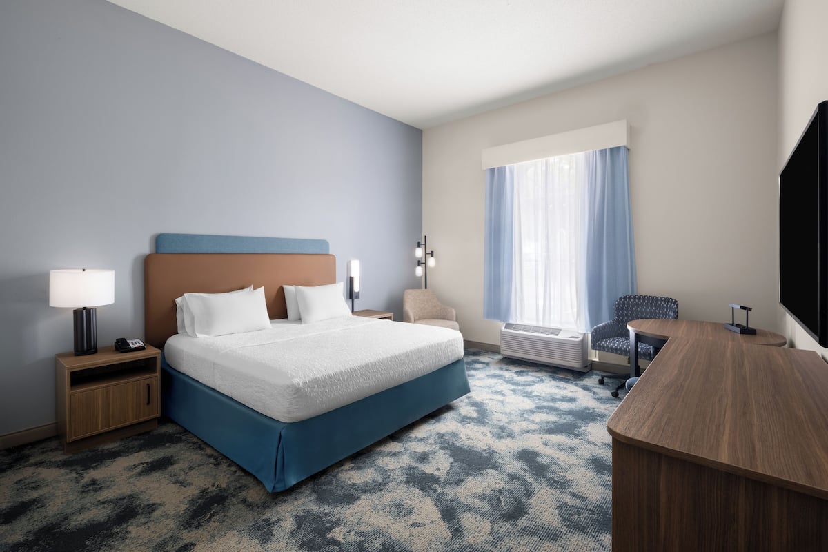 Quarto de hotel com uma cama grande, uma escrivaninha, uma cadeira, um abajur, uma mesa de cabeceira e uma janela com cortinas.
