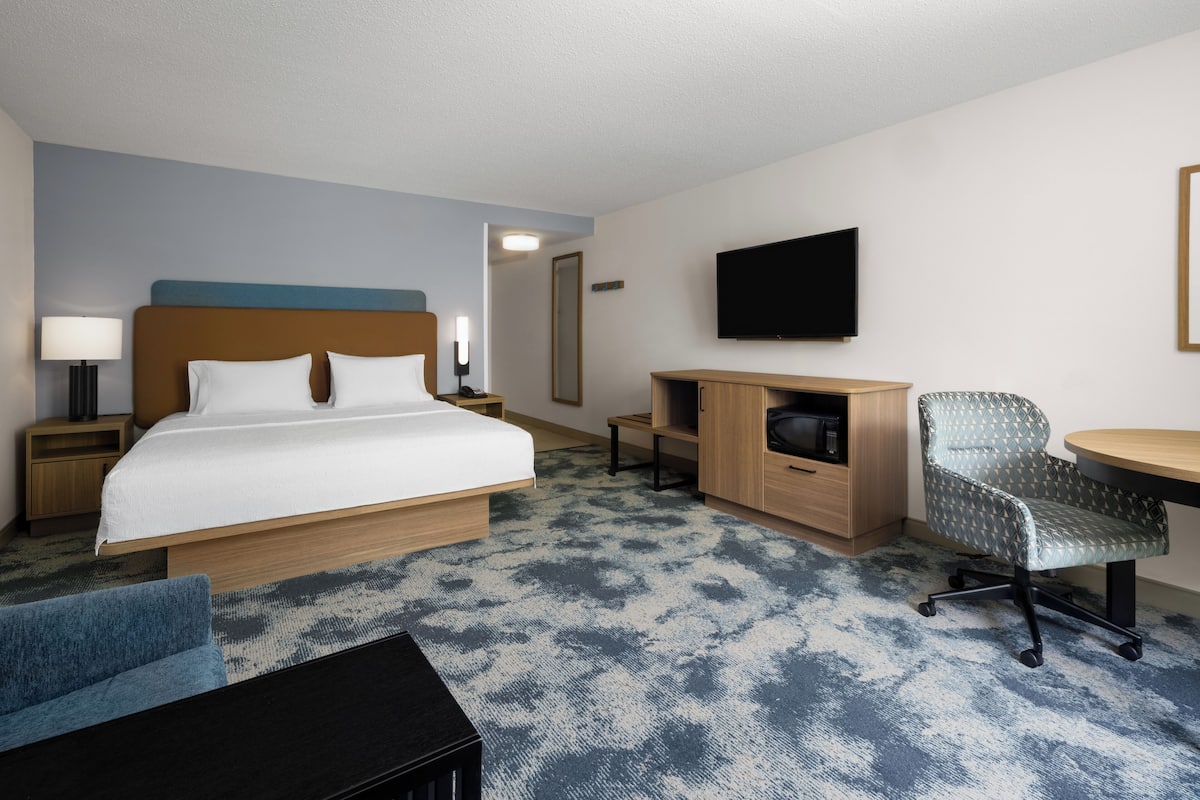 Quarto de hotel com cama, escrivaninha, cadeira, televisão e luminária.