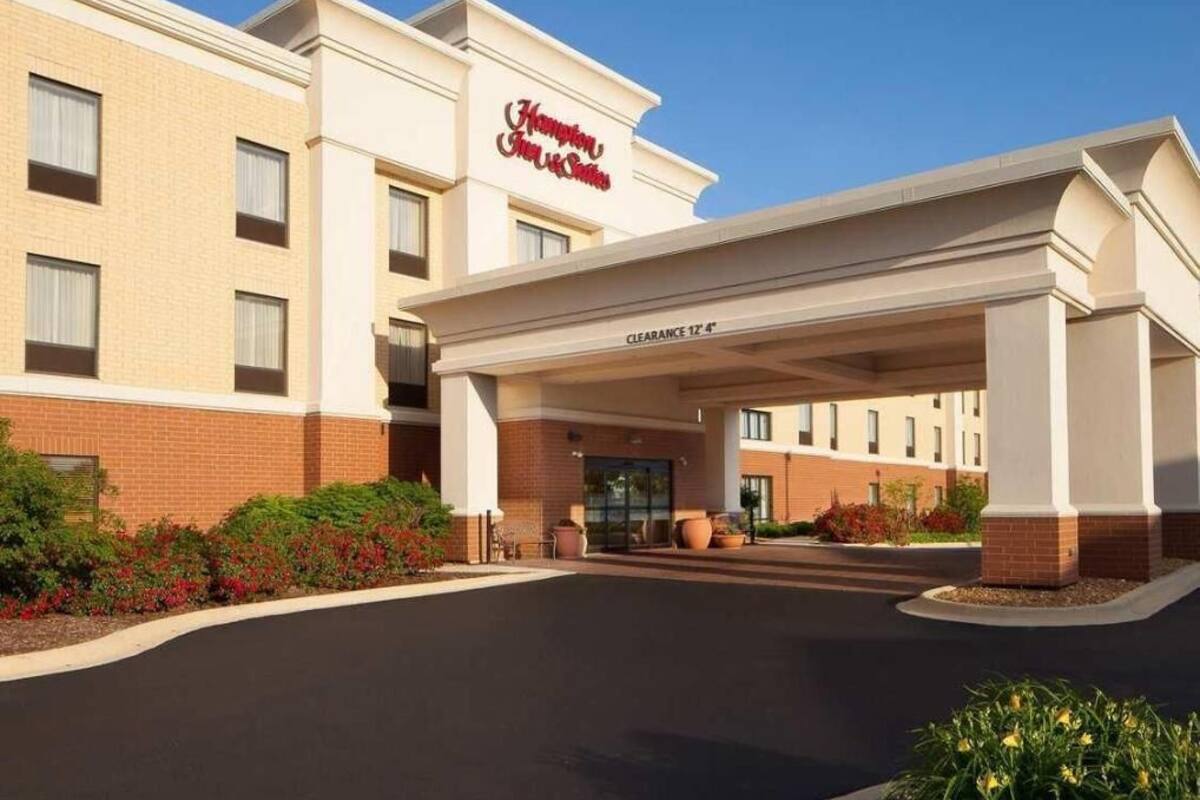 Entrada de um hotel Hampton Inn & Suites, com acesso por uma entrada livre, área de espera coberta e fachada de tijolos.