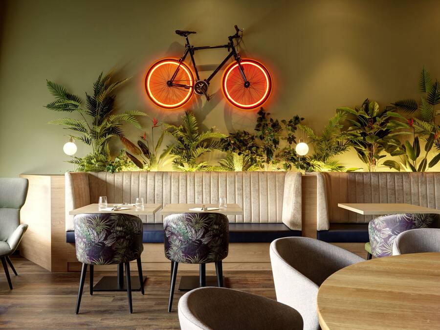Interior de restaurante com área de estar, incluindo um banco e cadeiras, uma bicicleta decorativa na parede e diversos vasos de plantas.