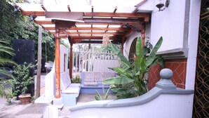 Uma área de pátio coberta com pérgola de madeira, parede branca com padrão de azulejos decorativos e um vaso com planta.