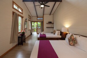 Quarto de hotel com duas camas, um ventilador de teto, janela com cortinas, uma mesa pequena com um vaso e vista para o exterior.