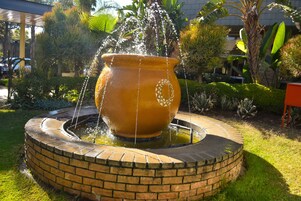 Uma grande fonte circular de tijolos com jatos de água, cercada por vegetação exuberante e um edifício de tijolos ao fundo.