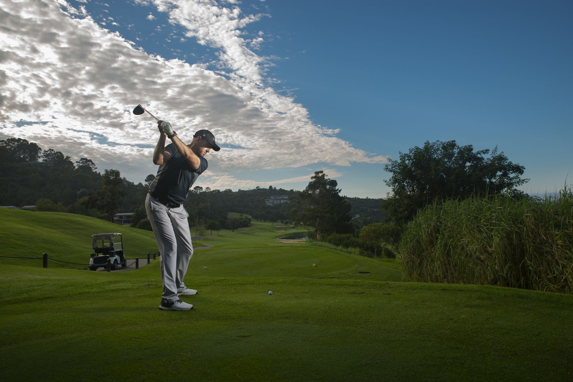 Um campo de golfe com um jogador a meio do movimento, um carrinho de golfe e um céu claro.