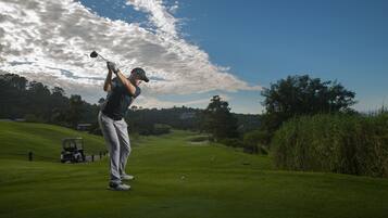 Um campo de golfe com um jogador a meio do movimento, um carrinho de golfe e um céu claro.