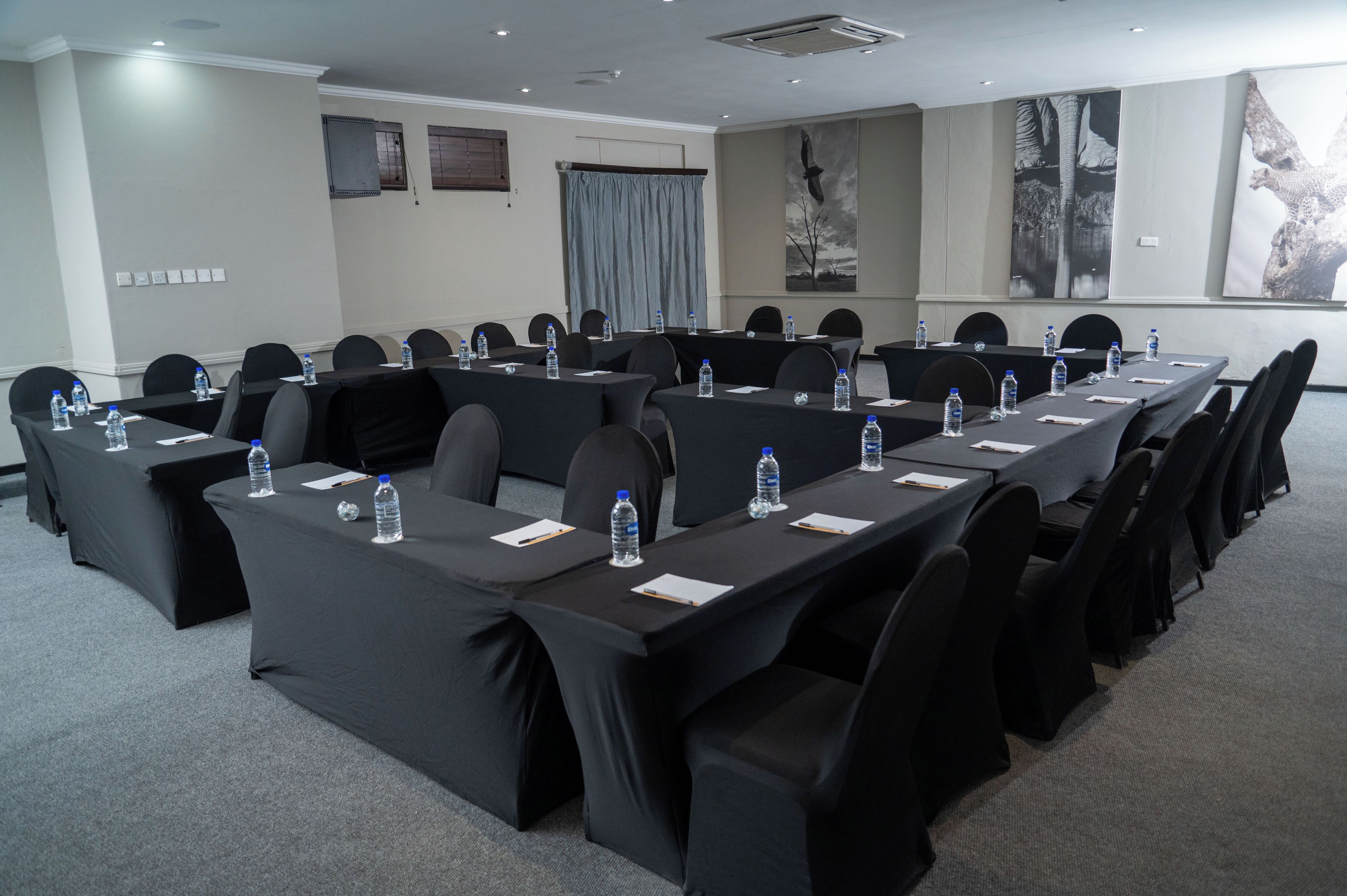 Sala de conferências com mesas compridas, cadeiras, garrafas de água e uma tela de projeção.