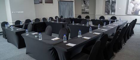 Sala de conferências com mesas compridas, cadeiras, garrafas de água e uma tela de projeção.