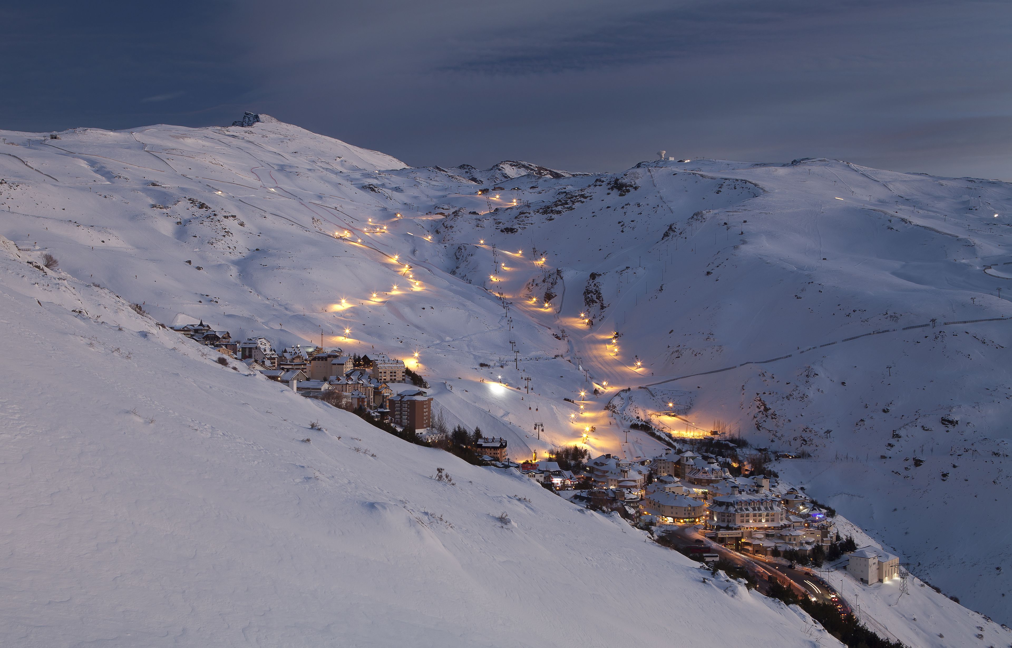 Montanha coberta de neve com uma cidade na base, apresentando pistas de esqui iluminadas e edifícios.