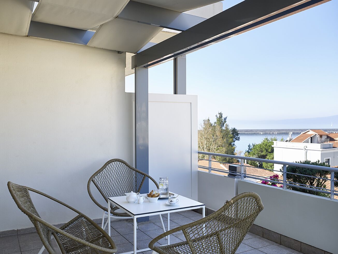 Varanda com cadeiras de vime, uma mesa pequena e vista para a água e edifícios.
