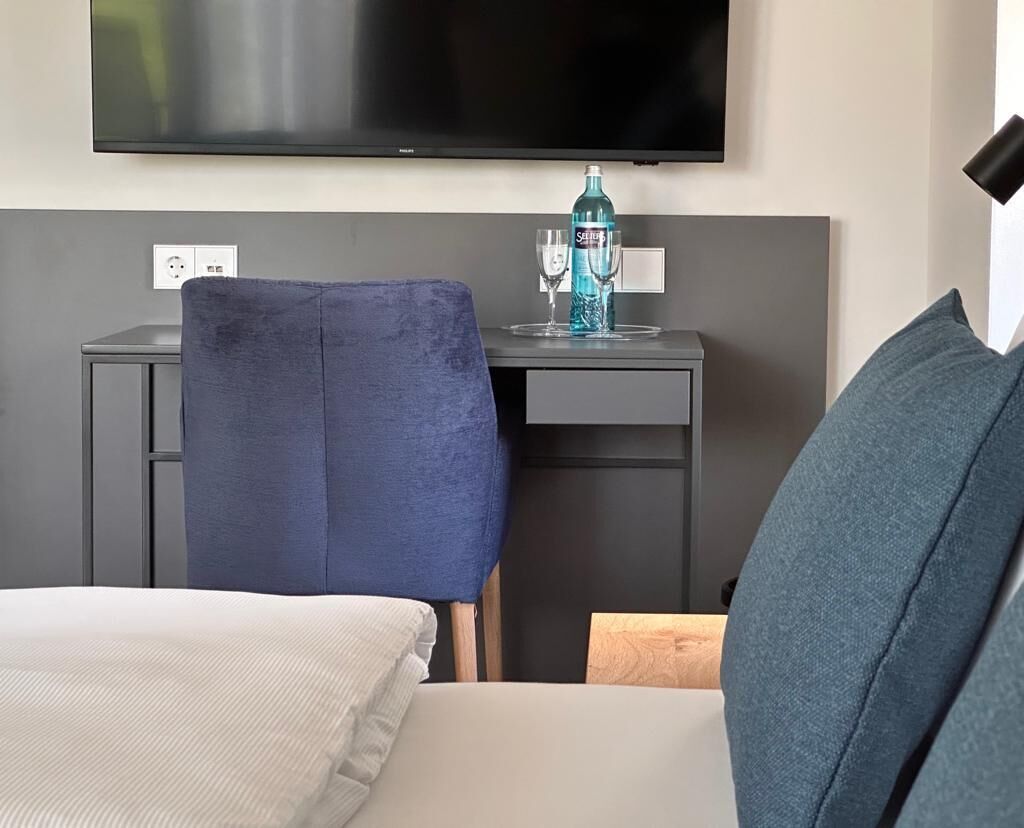 Quarto de hotel com cama, escrivaninha, televisão fixada na parede e uma garrafa de água sobre a escrivaninha.