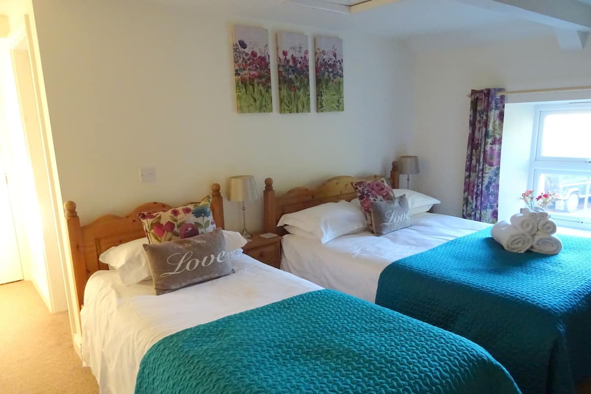Quarto de hotel com duas camas, cada uma com colcha e travesseiros azuis-turquesa, cabeceira de madeira e uma janela com cortinas.