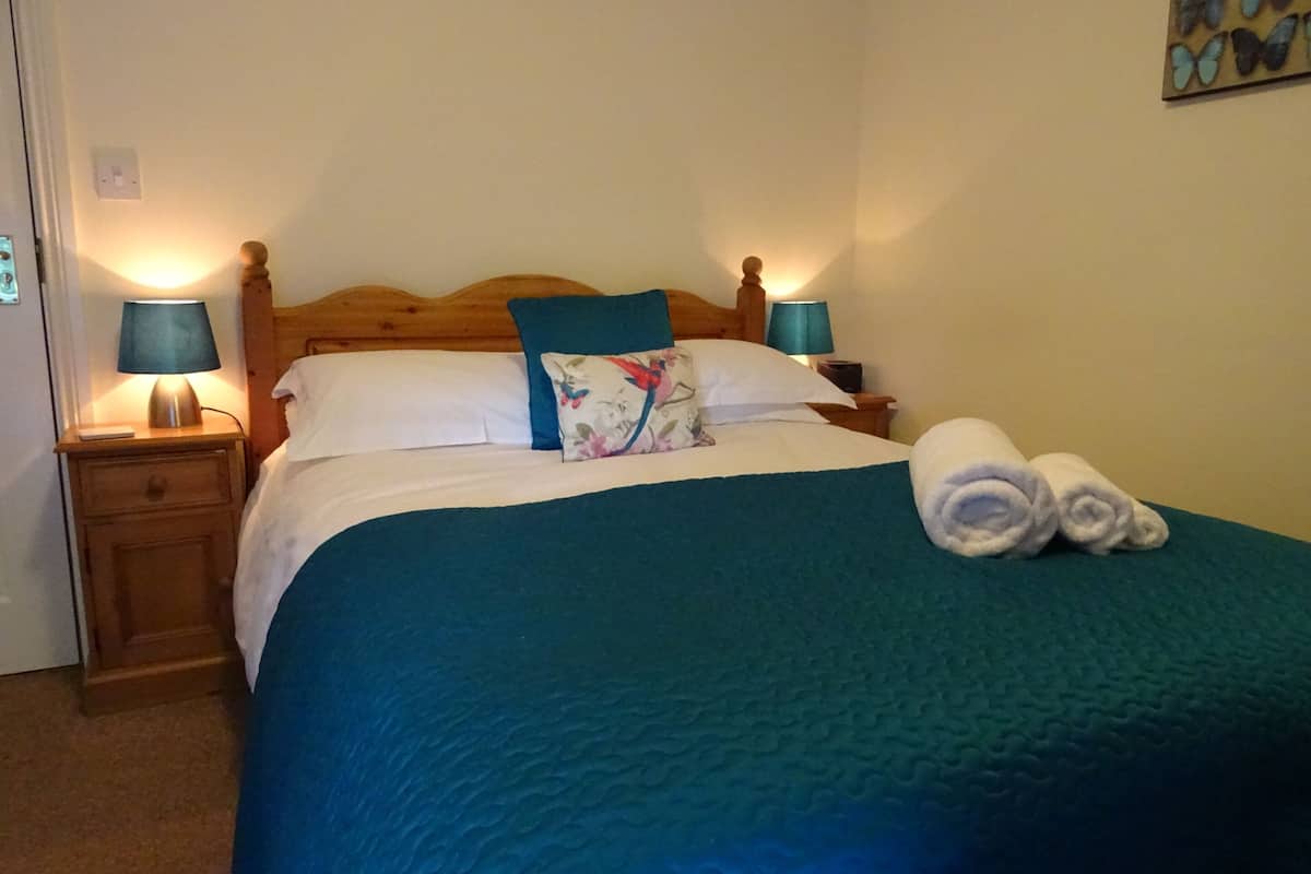 Cama bem arrumada com um edredom azul, dois travesseiros e um travesseiro floral. Duas luminárias de cabeceira estão sobre mesas de cabeceira de madeira, e um par de toalhas enroladas na cama.