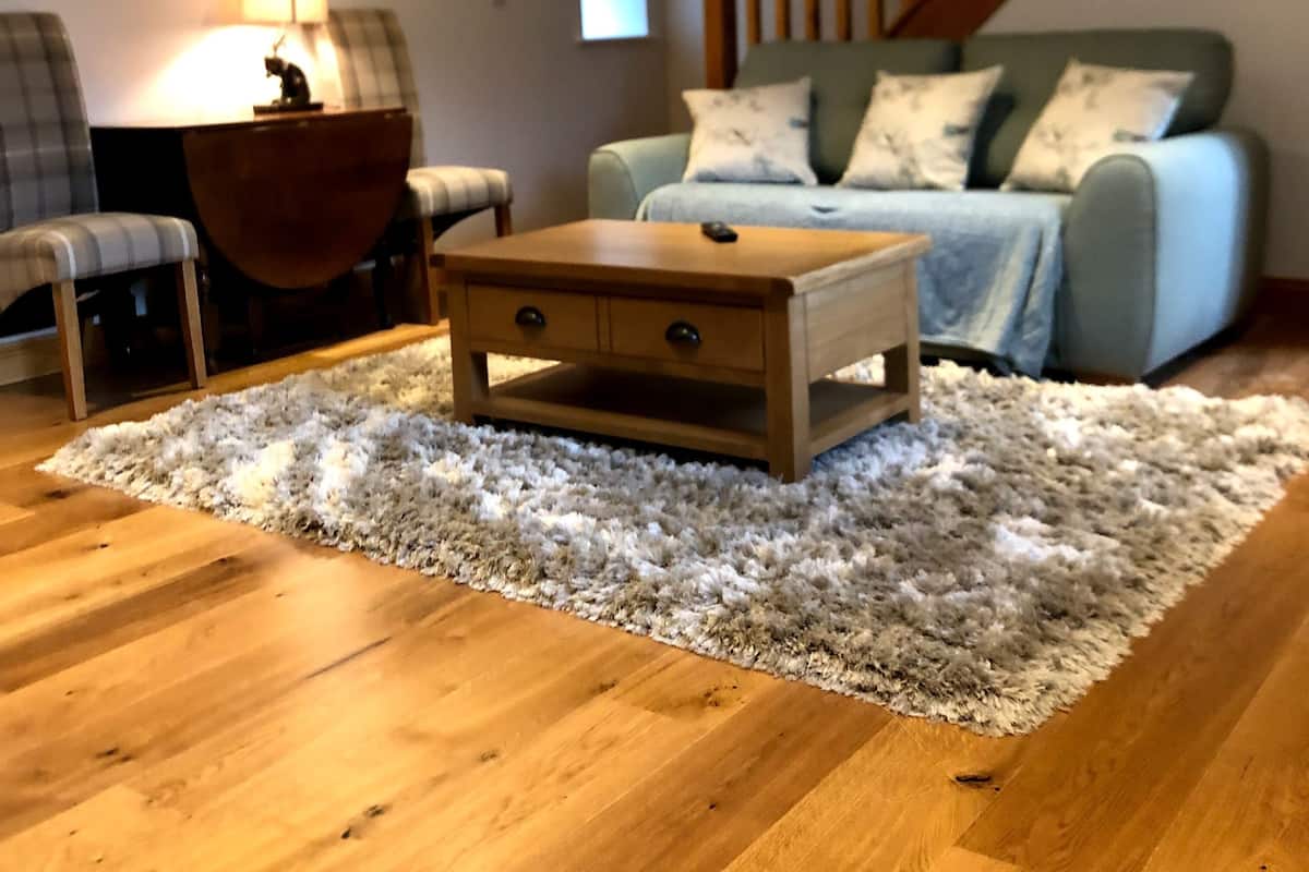 Uma sala de estar com uma mesa de centro de madeira, um sofá cinza com almofadas estampadas, uma poltrona xadrez, um abajur e uma escada ao fundo.