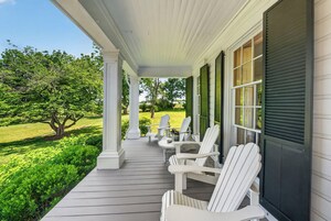 Varanda coberta com cadeiras Adirondack brancas, uma mesa pequena e vista para uma paisagem verde.