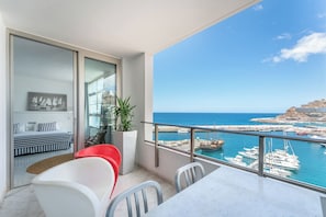 Uma varanda com vista para uma marina e o mar, mesa de jantar, cadeiras, sofá e uma cama com roupa de cama listrada.
