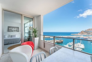 Uma varanda com vista para uma marina e o mar, mesa de jantar, cadeiras, sofá e uma cama com roupa de cama listrada.