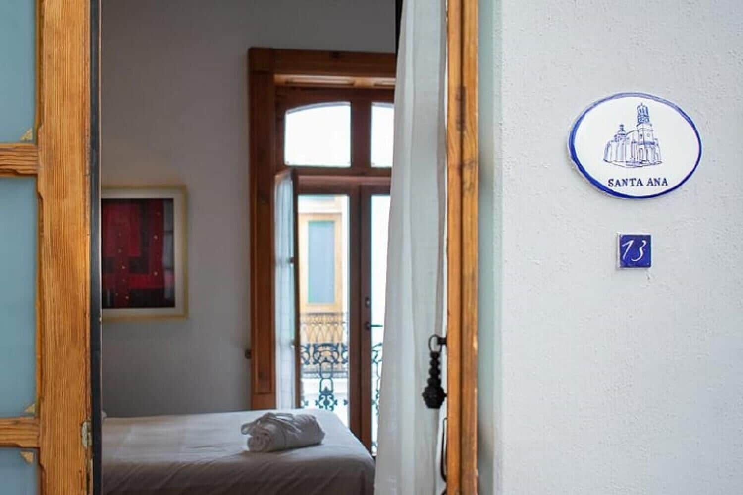 Quarto de hotel com cama, janela e porta que d á para uma varanda. Há uma placa na parede com o nome do hotel e o número do quarto.