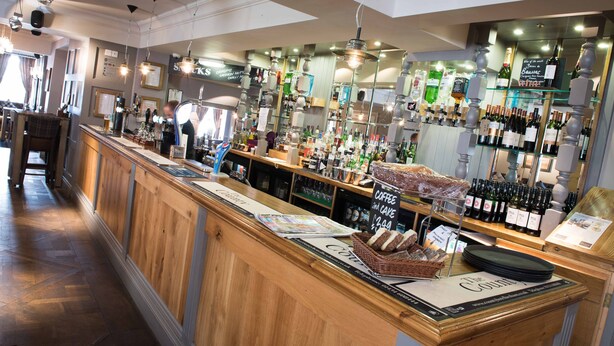 Uma área de bar com balcão de madeira, prateleiras com diversos frascos e um letreiro de café e bolo.