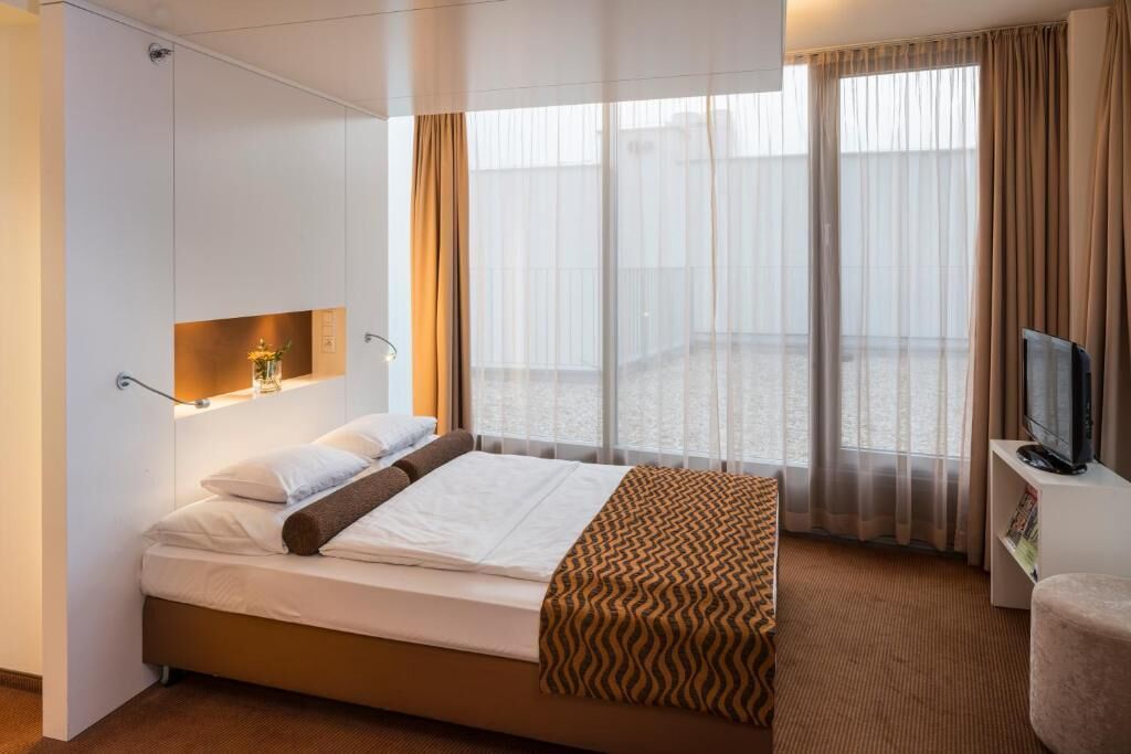 Quarto de hotel com cama, televisão em suporte, janela com cortinas e vista para varanda.