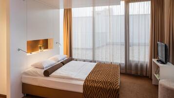 Quarto de hotel com cama, televisão em suporte, janela com cortinas e vista para varanda.