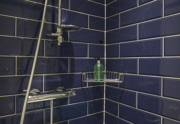 Um banheiro com azulejos azul escuro, chuveiro cromado e um dosador de sabonete em uma prateleira.