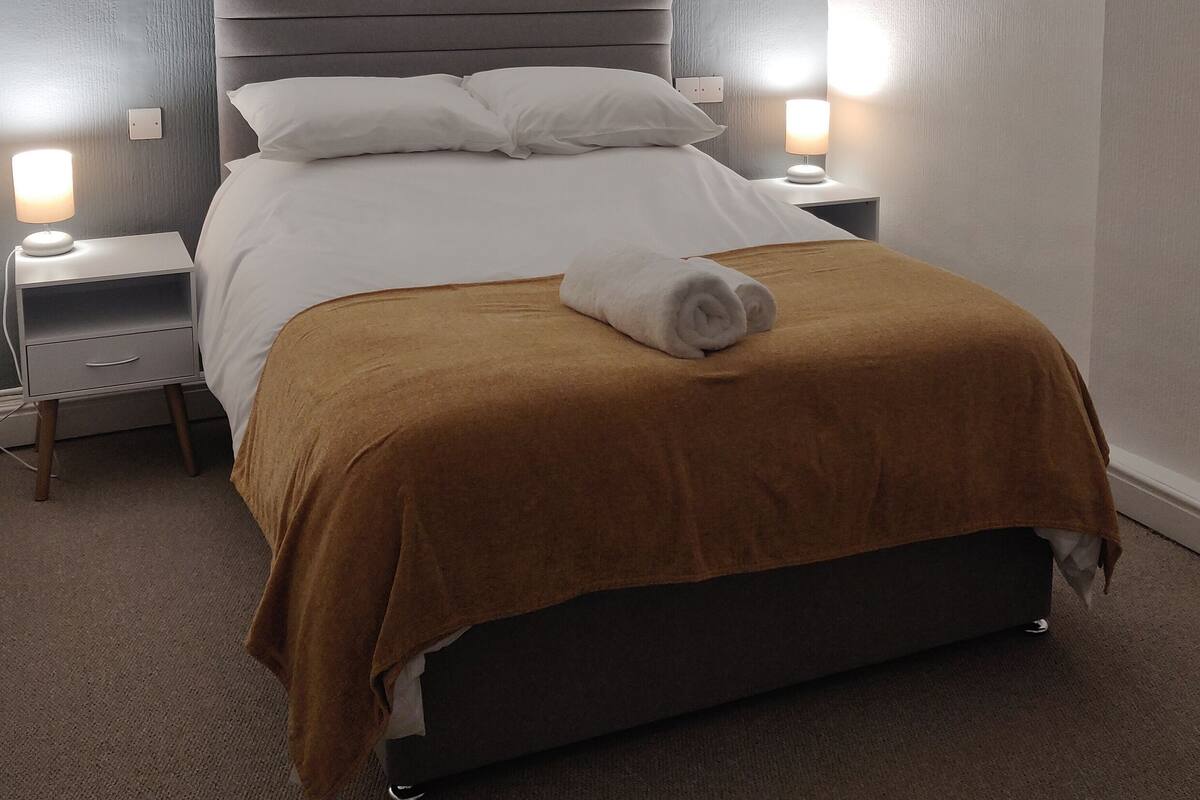 Uma cama bem arrumada com um cobertor marrom, duas mesas de cabeceira com abajures e uma cabeceira em frente a uma parede lisa.