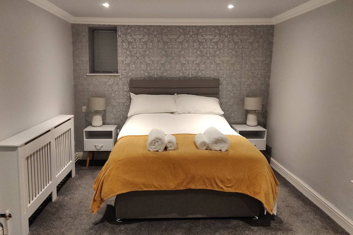 Quarto com cama, duas mesas de cabeceira, um criado-mudo, radiador e janela com persianas.