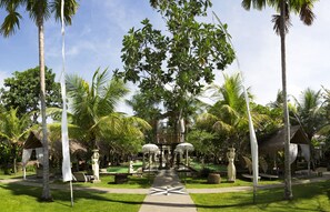 Um resort tropical com um caminho central que leva a um gazebo, cercado por palmeiras e estruturas com telhados de palha.