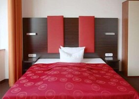 Quarto de hotel com uma cama, uma mesinha de cabeceira, um edredom vermelho e branco estampado e uma cabeceira de madeira com dois painéis vermelhos.