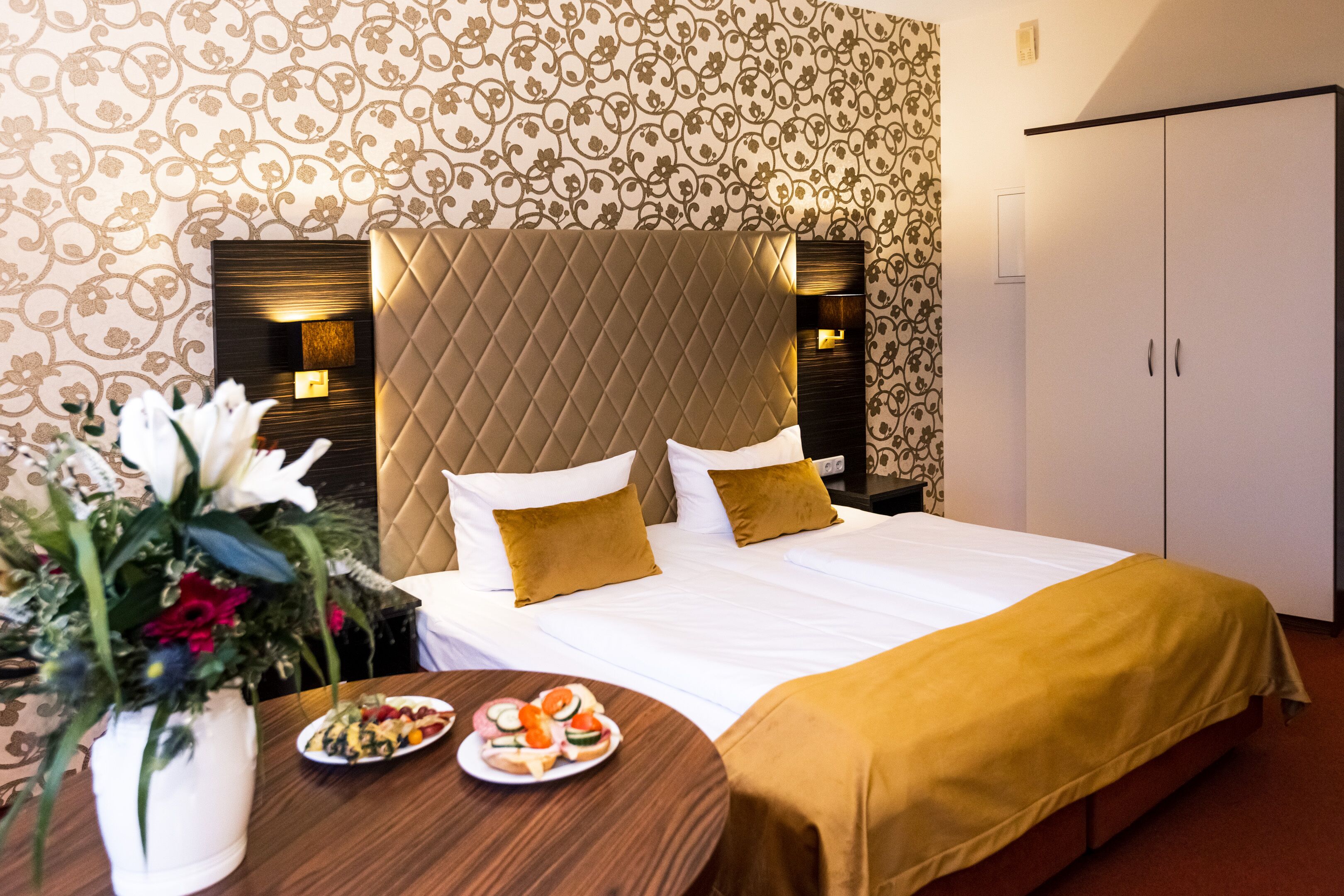 Quarto de hotel com uma cama grande, uma mesinha de cabeceira com um vaso de flores e uma travessa de frutas.
