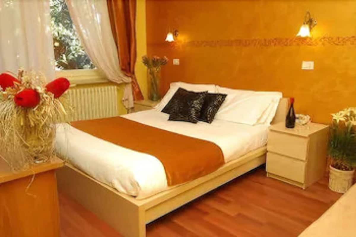 Quarto com cama de madeira, roupa de cama branca, mesa de cabeceira, um vaso com flores e janela com cortinas.