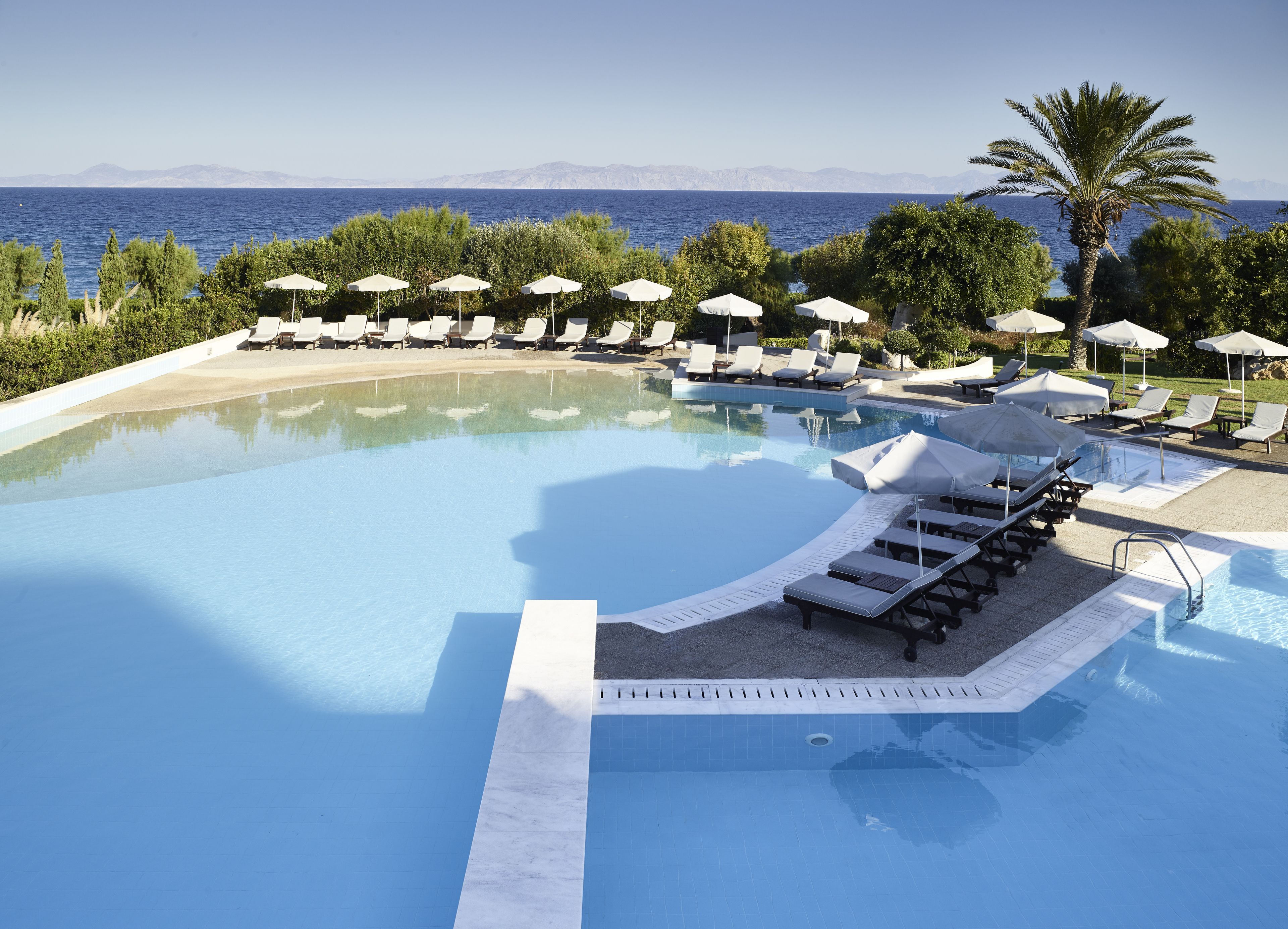 Piscina com espreguiçadeiras e guarda-sóis, cercada por vegetação e vista para o mar.